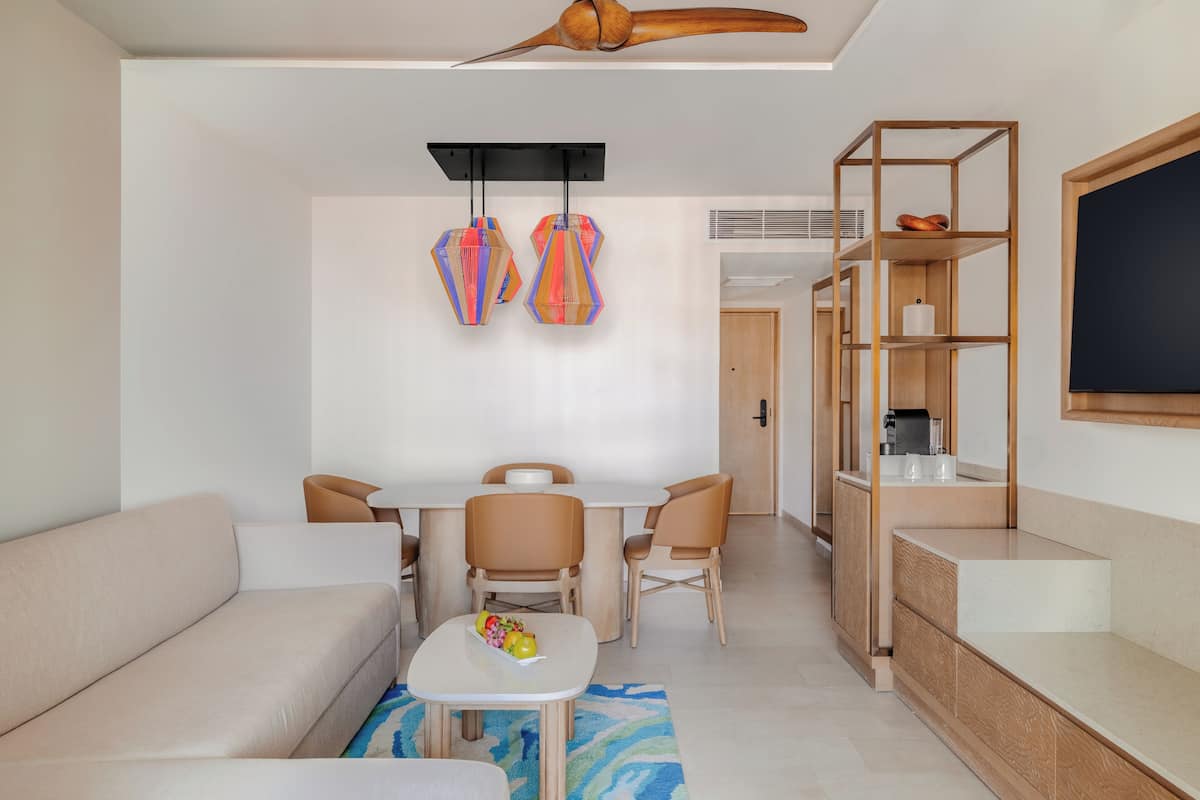 Uma sala de estar moderna com um sofá, uma mesa de jantar com cadeiras, uma pequena mesa de centro com frutas, uma luminária pendente colorida e uma estante de madeira.
