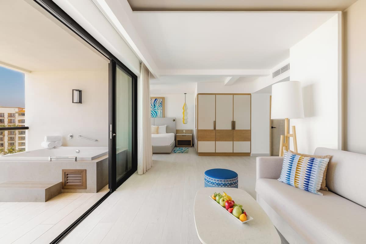 Quarto de hotel moderno com varanda, sofá, mesa de jantar com frutas, cama e guarda-roupa.