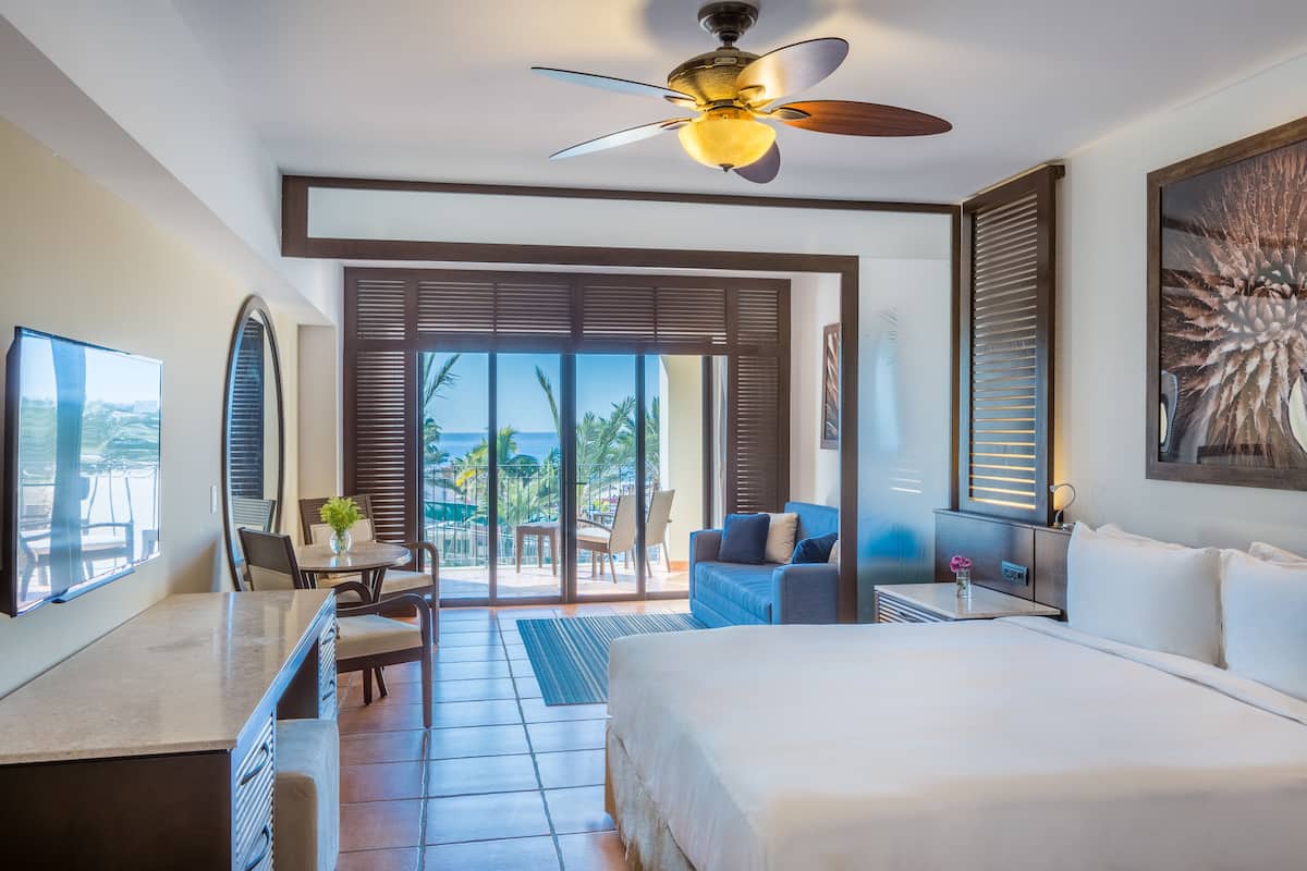 Quarto de hotel com cama, escrivaninha, poltrona, sofá, ventoinha no teto e uma grande janela com vista para palmeiras.