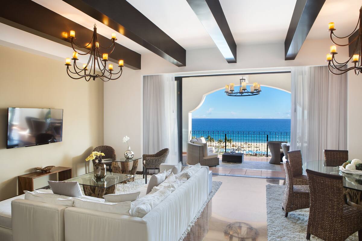 Uma sala de estar ampla, com um sofá grande, uma mesa de centro de vidro e uma TV fixada na parede. Há vista para o oceano através de uma grande janela.