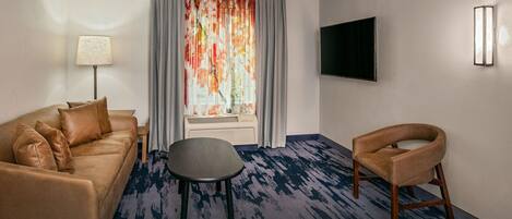 Quarto de hotel com sofá, uma cadeira, uma mesinha, televisão de tela plana e uma janela com cortinas.