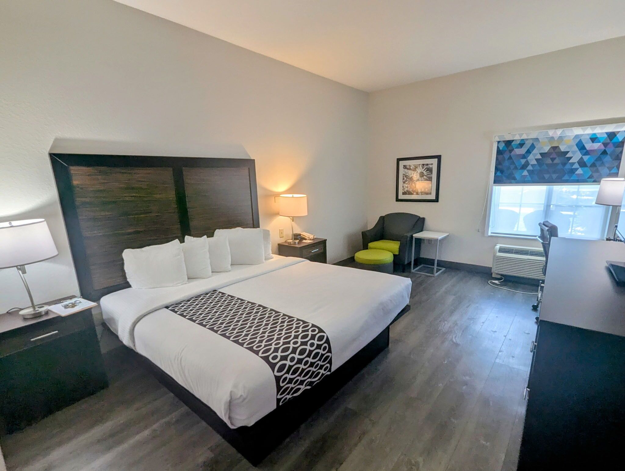 Quarto de hotel com uma cama grande, uma mesinha de cabeceira com abajur, uma cadeira com almofada verde, uma mesa pequena e uma janela com cortina geométrica.