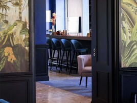 Uma área de bar com banquetas altas e uma cadeira estofada, um vaso com plantas secas e uma janela grande ao fundo.