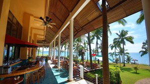 Uma área de bar ao ar livre coberta, com mesas e cadeiras, com vista para uma praia com palmeiras e o oceano.