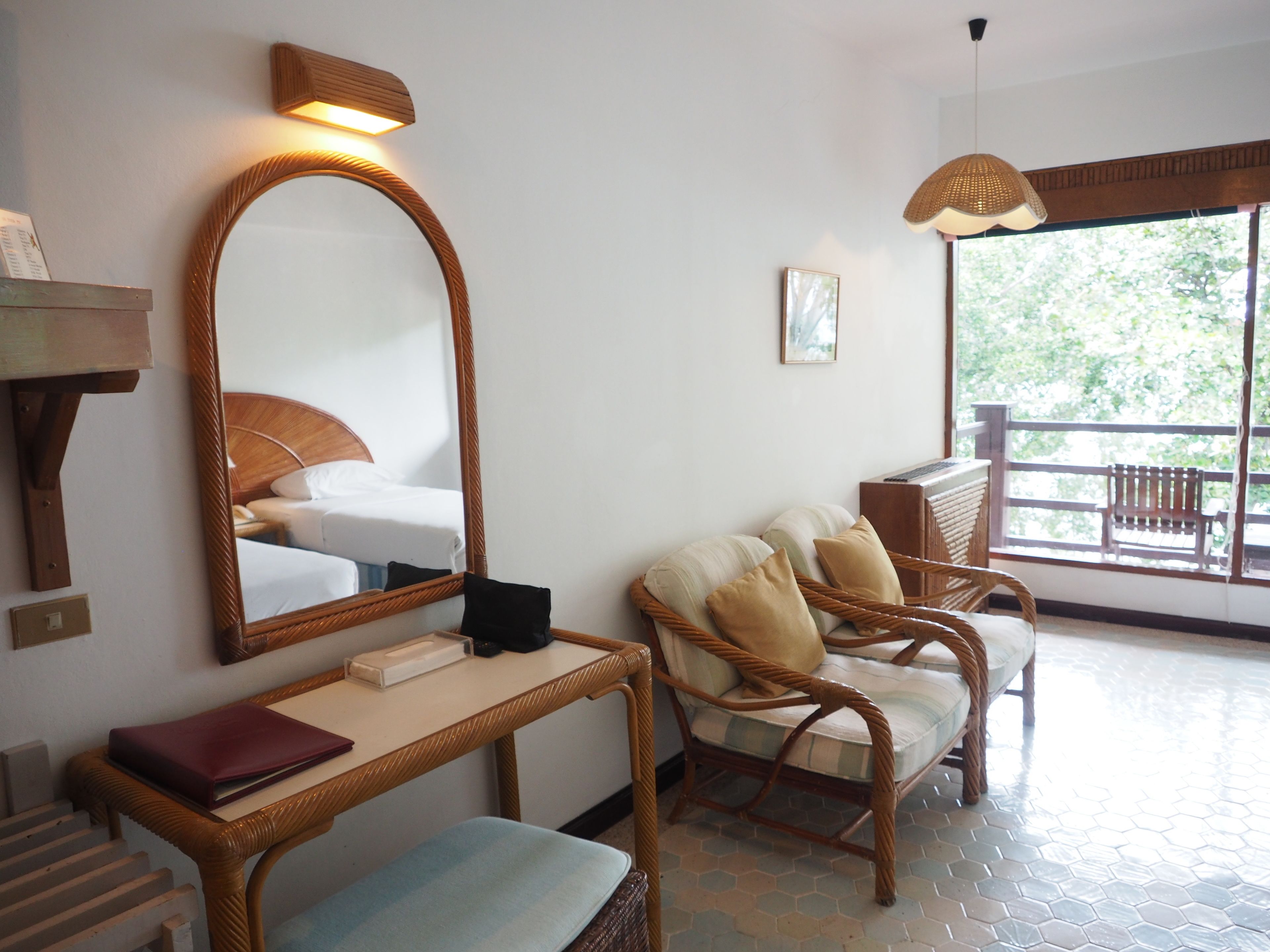 Quarto de hotel com uma cama, um espelho grande, uma escrivaninha com cadeira, um sofá de vime e vista para o exterior.
