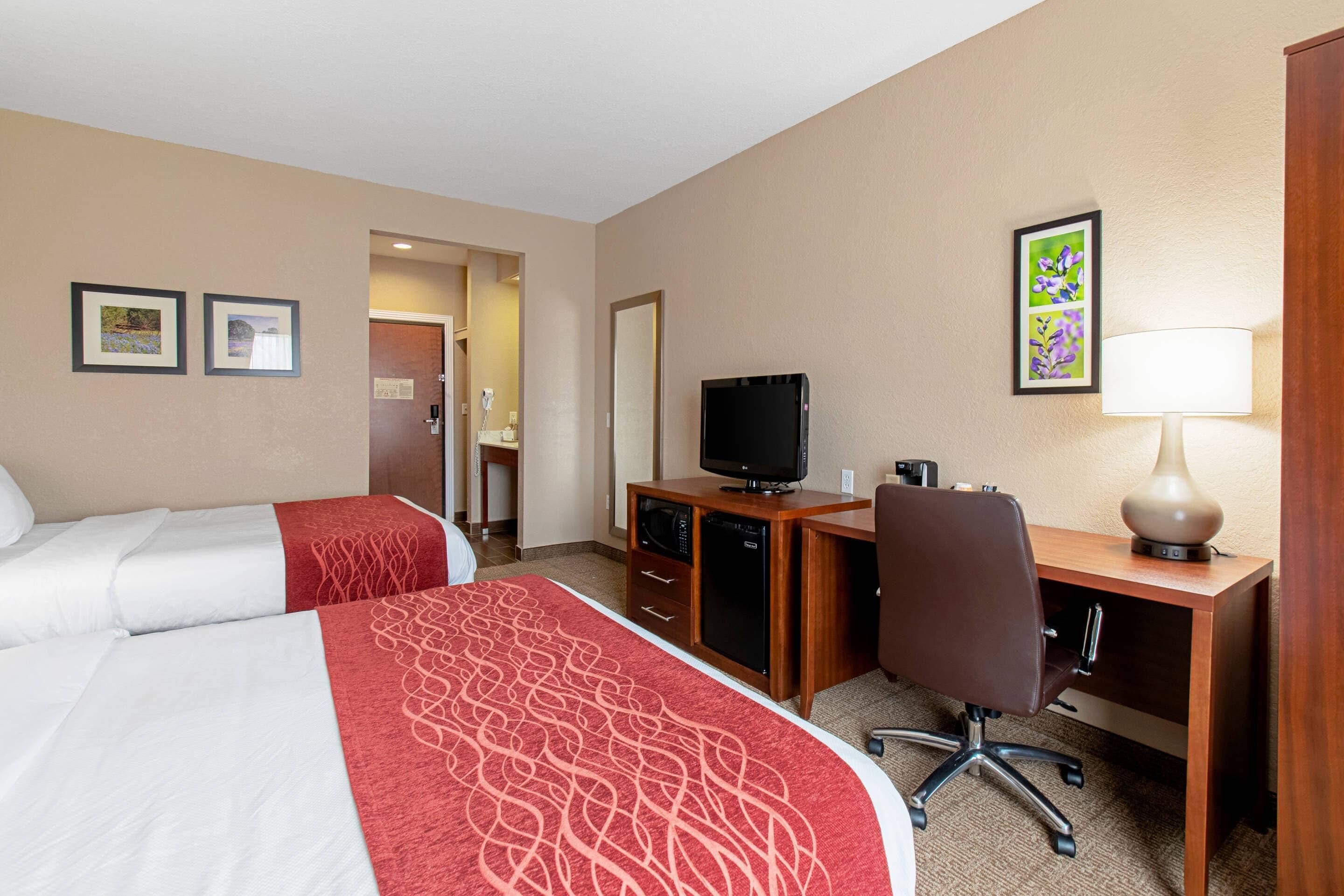 Quarto de hotel com duas camas, uma escrivaninha com computador, televisão e luminária.