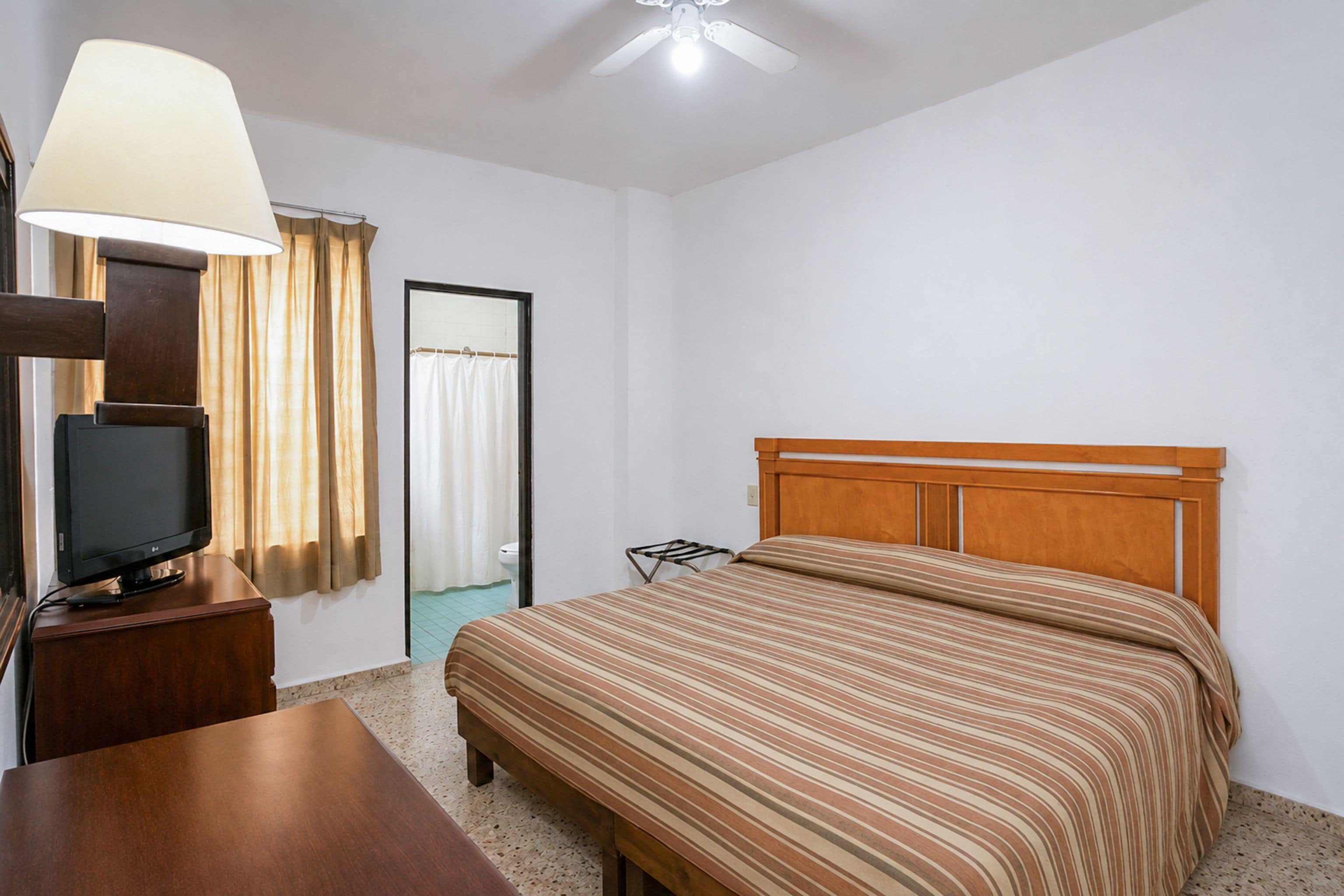 Quarto de hotel com cama, escrivaninha com televisão, ventoinha no teto e porta para o banheiro.