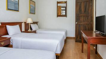 Quarto de hotel com duas camas, uma mesa de cabeceira com abajur, porta de madeira e televisão sobre suporte de madeira.