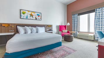 Quarto de hotel com uma cama grande, uma mesinha de cabeceira, uma cadeira, uma janela com cortinas e uma obra de arte na parede.