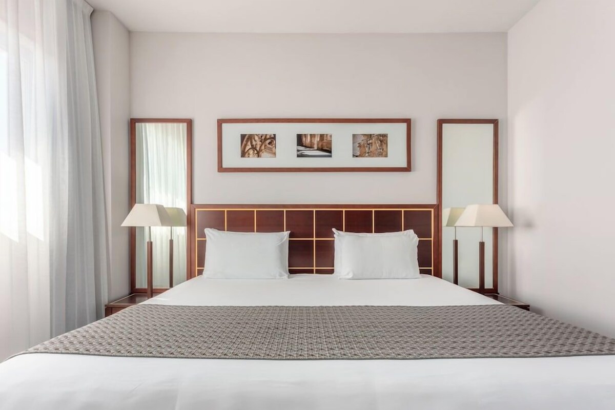 Quarto de hotel com uma cama grande, duas luminárias de cabeceira, um painel de cabeceira com padrão de grade e três quadros pendurados na parede.