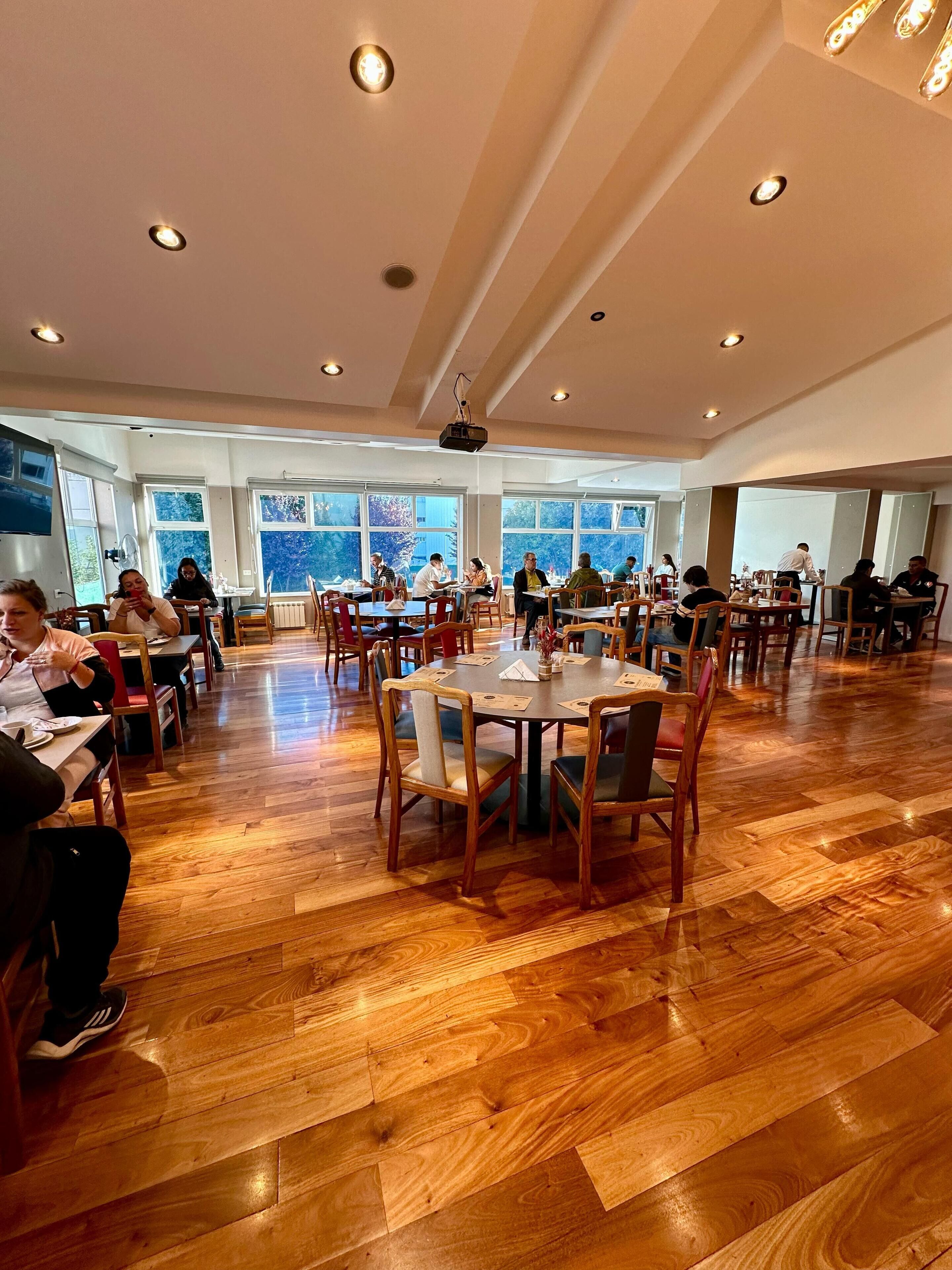 Ampla área de refeições com piso de madeira e janelas grandes. As mesas estão dispostas em formato de U, e há várias luminárias no teto.