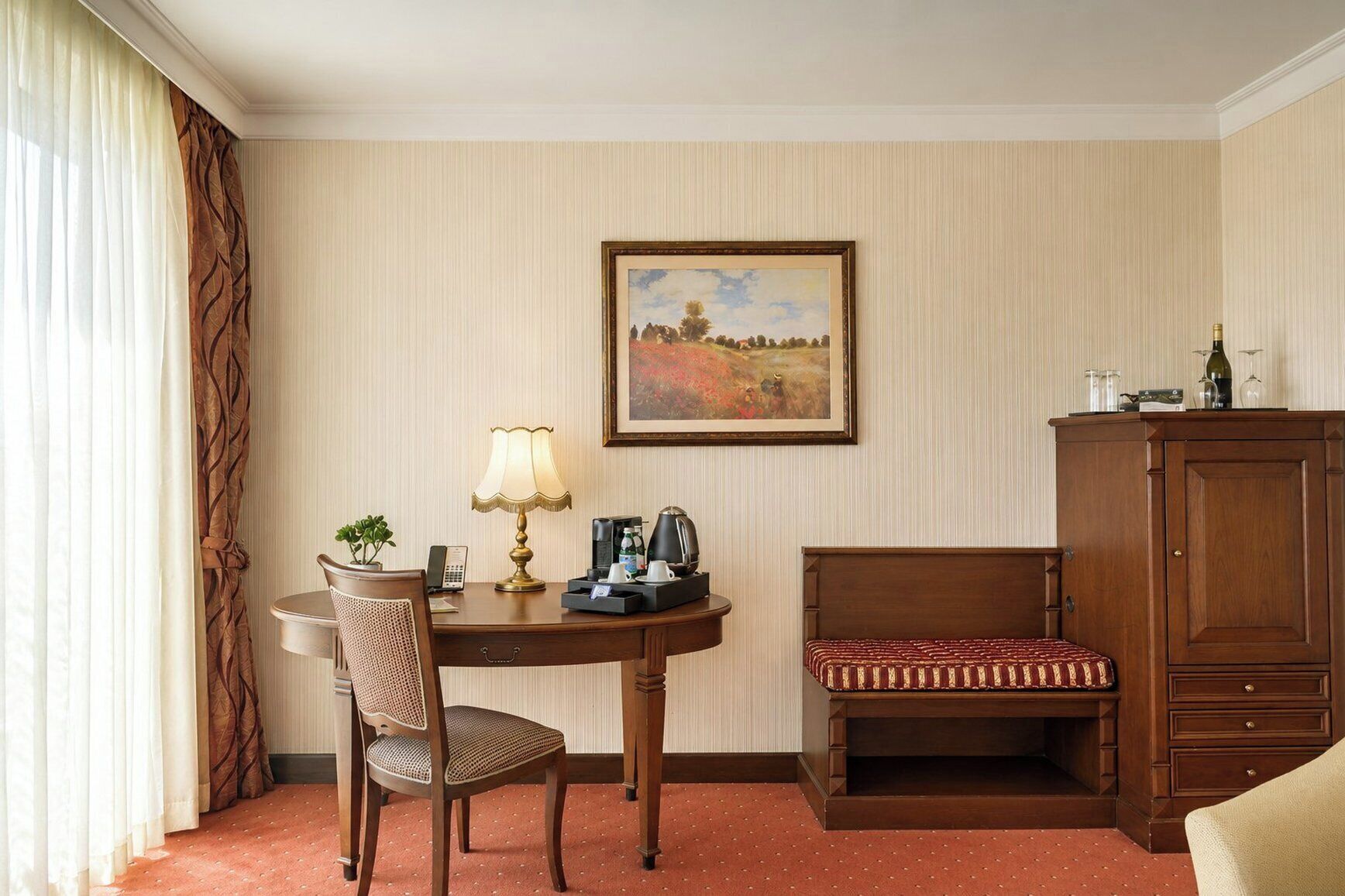Quarto de hotel com uma mesa de jantar, uma cadeira, um abajur, um quadro na parede, um armário e uma janela com cortinas.