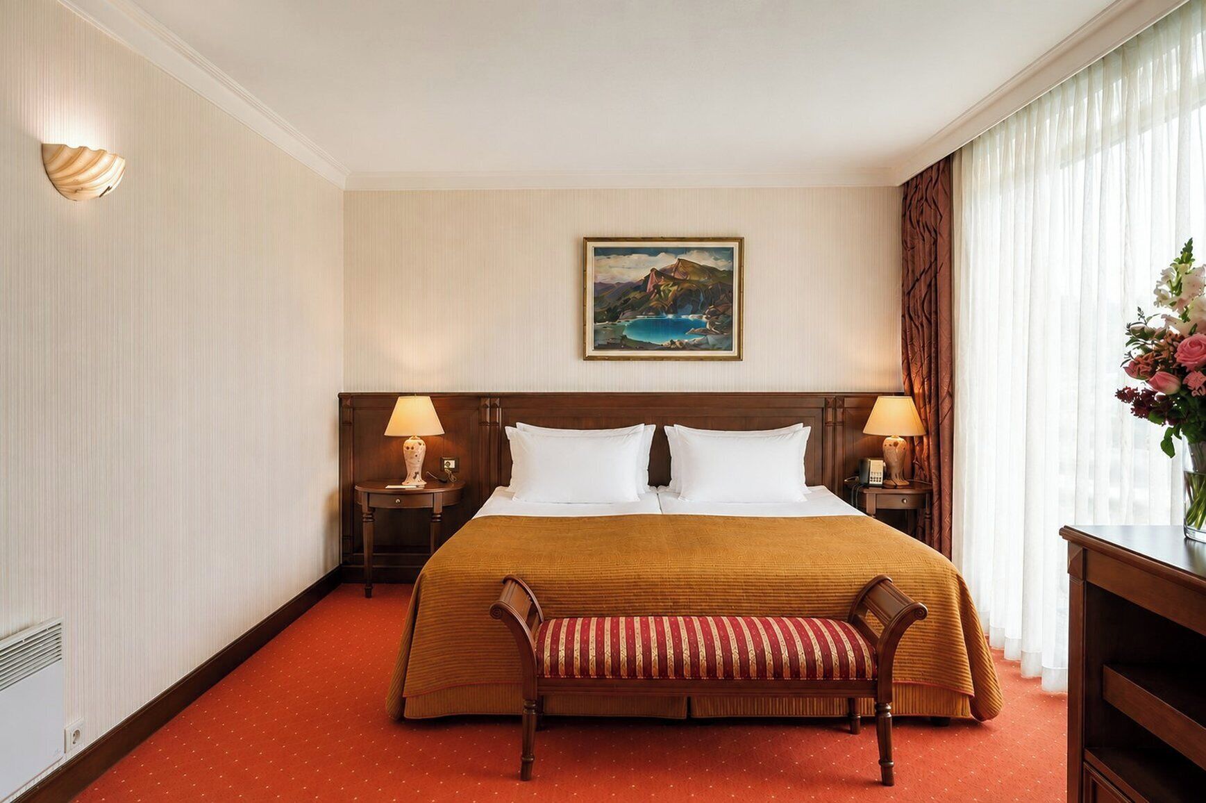 Quarto de hotel com uma cama grande, duas mesas de cabeceira com abajures, um banco, um quadro na parede e uma janela com cortinas.