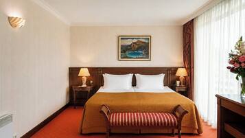 Quarto de hotel com uma cama grande, duas mesas de cabeceira com abajures, um banco, um quadro na parede e uma janela com cortinas.