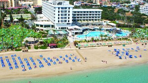 Vista aérea de um resort com uma piscina grande, várias áreas de praia com espreguiçadeiras e um edifício de hotel ao fundo.