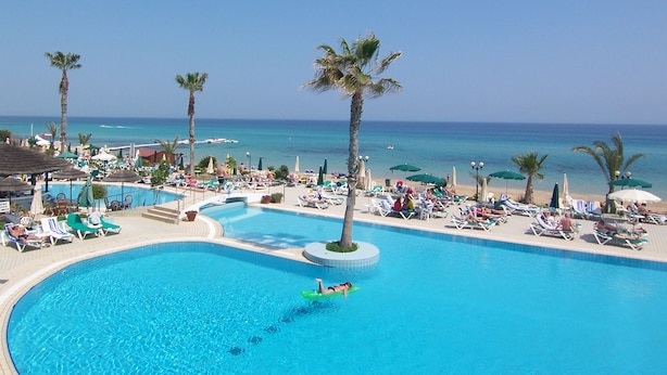Uma área de piscina com espreguiçadeiras e palmeiras, com vista para uma praia.