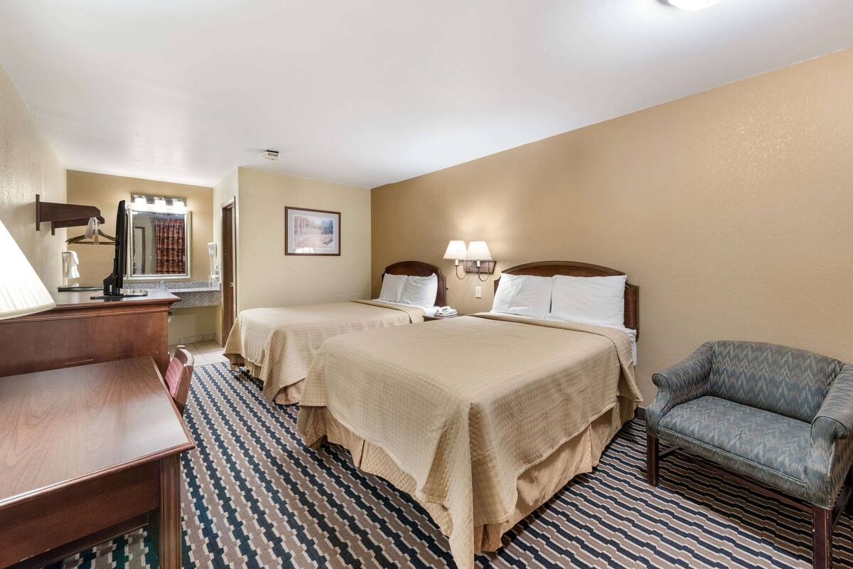 Quarto de hotel com duas camas, uma escrivaninha, uma cadeira e um tapete estampado.