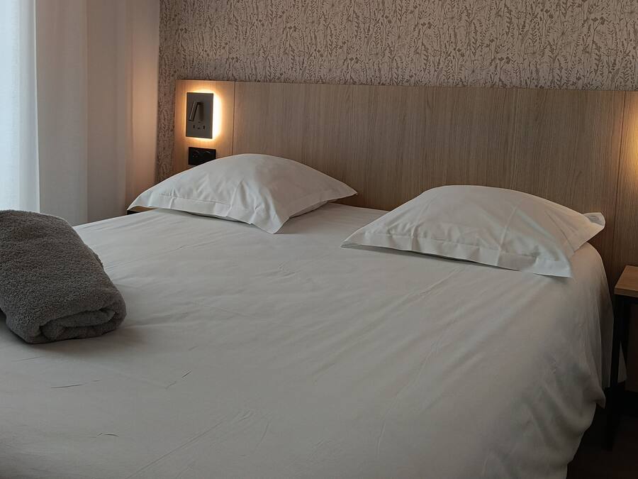 Cama bem arrumada com lençóis brancos e duas almofadas, uma toalha cinza e cabeceira de madeira com luminárias fixadas na parede.