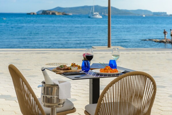 Mesa posta para dois, com uma garrafa de vinho tinto, uma taça de vinho branco, um prato de comida e uma garrafa de champanhe. A mesa fica em um pátio com vista para o mar e barcos ao fundo.