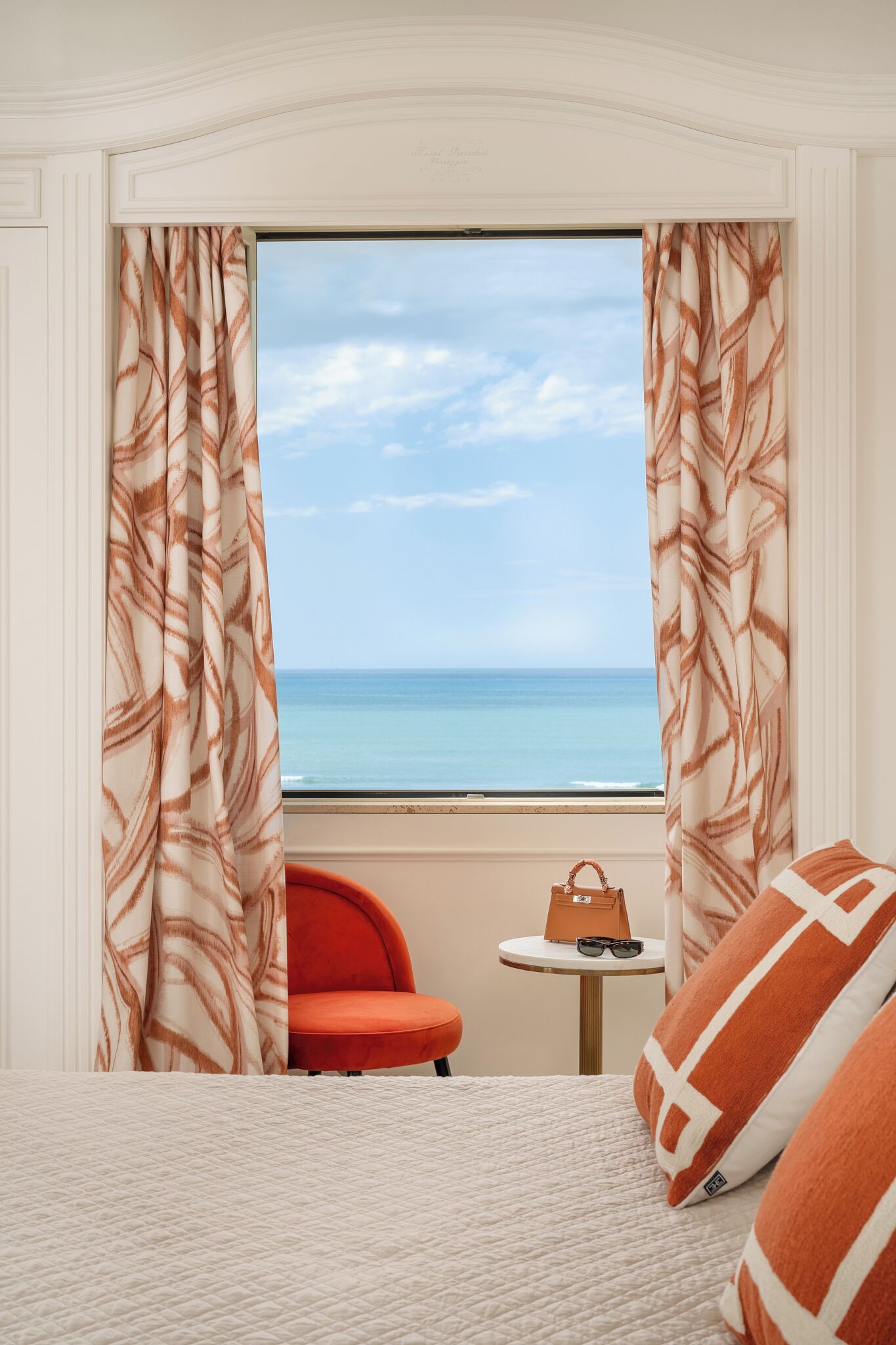 Quarto de hotel com cama, uma pequena mesa com uma bolsa, uma cadeira e vista para o oceano pelas cortinas.