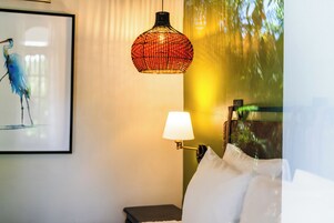 Quarto de hotel com cama, obra de arte emoldurada de um pássaro, luminária pendente vermelha e preta, e luminária fixada na parede.