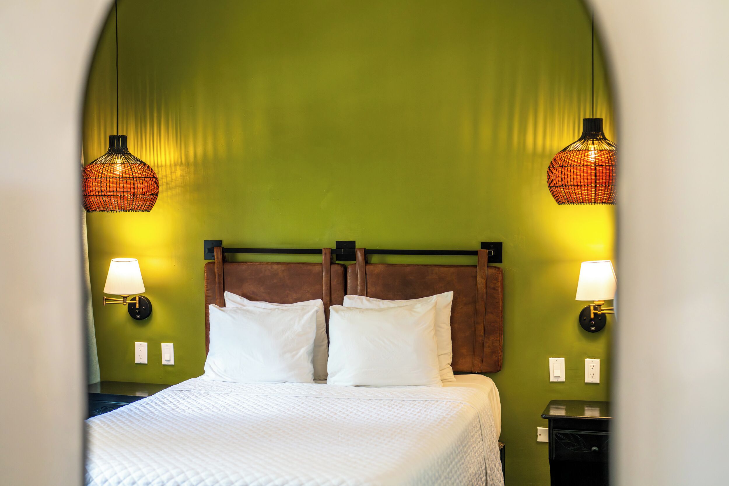 Quarto de hotel com parede verde, cabeceira de madeira, duas almofadas brancas e duas luminárias fixadas na parede.