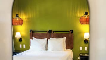Quarto de hotel com parede verde, cabeceira de madeira, duas almofadas brancas e duas luminárias fixadas na parede.
