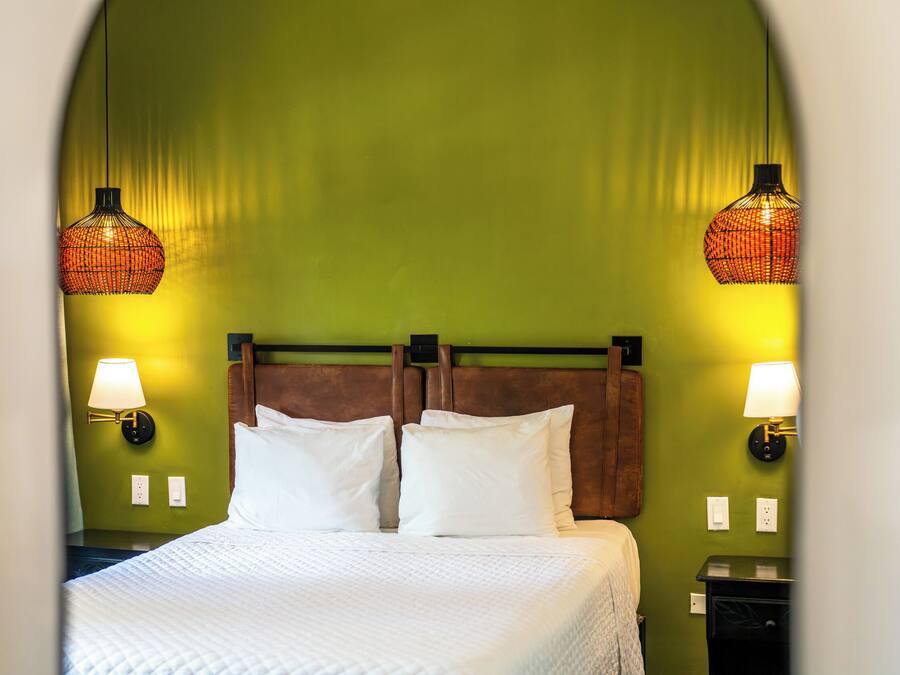 Quarto de hotel com parede verde, cabeceira de madeira, duas almofadas brancas e duas luminárias fixadas na parede.