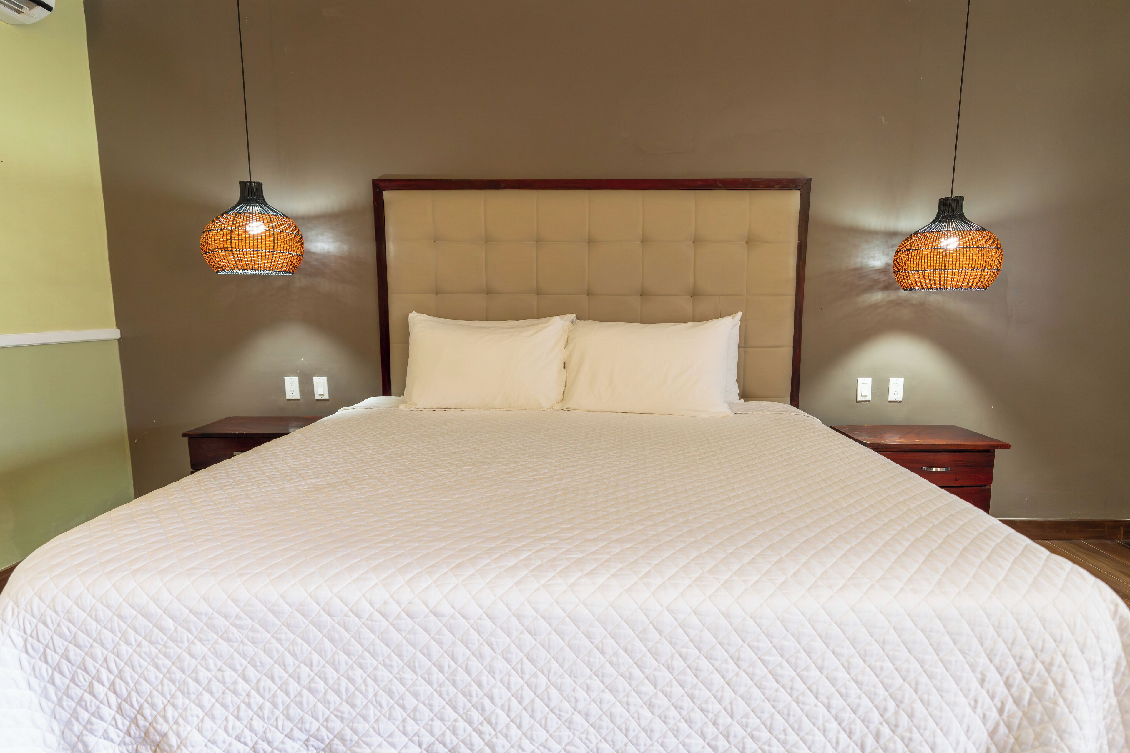Cama bem arrumada com edredom branco acolchoado, duas mesas de cabeceira e duas luminárias pendentes com design trançado.