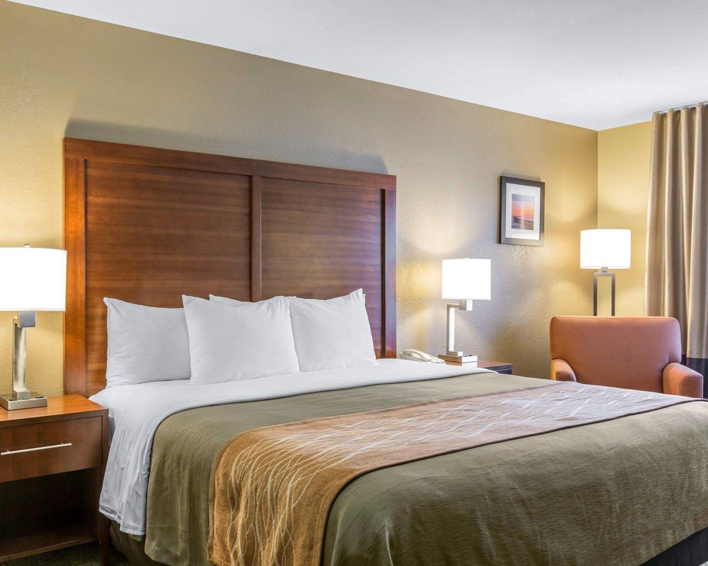 Quarto de hotel com uma cama grande, duas mesas de cabeceira com abajures, uma cadeira e um quadro na parede.