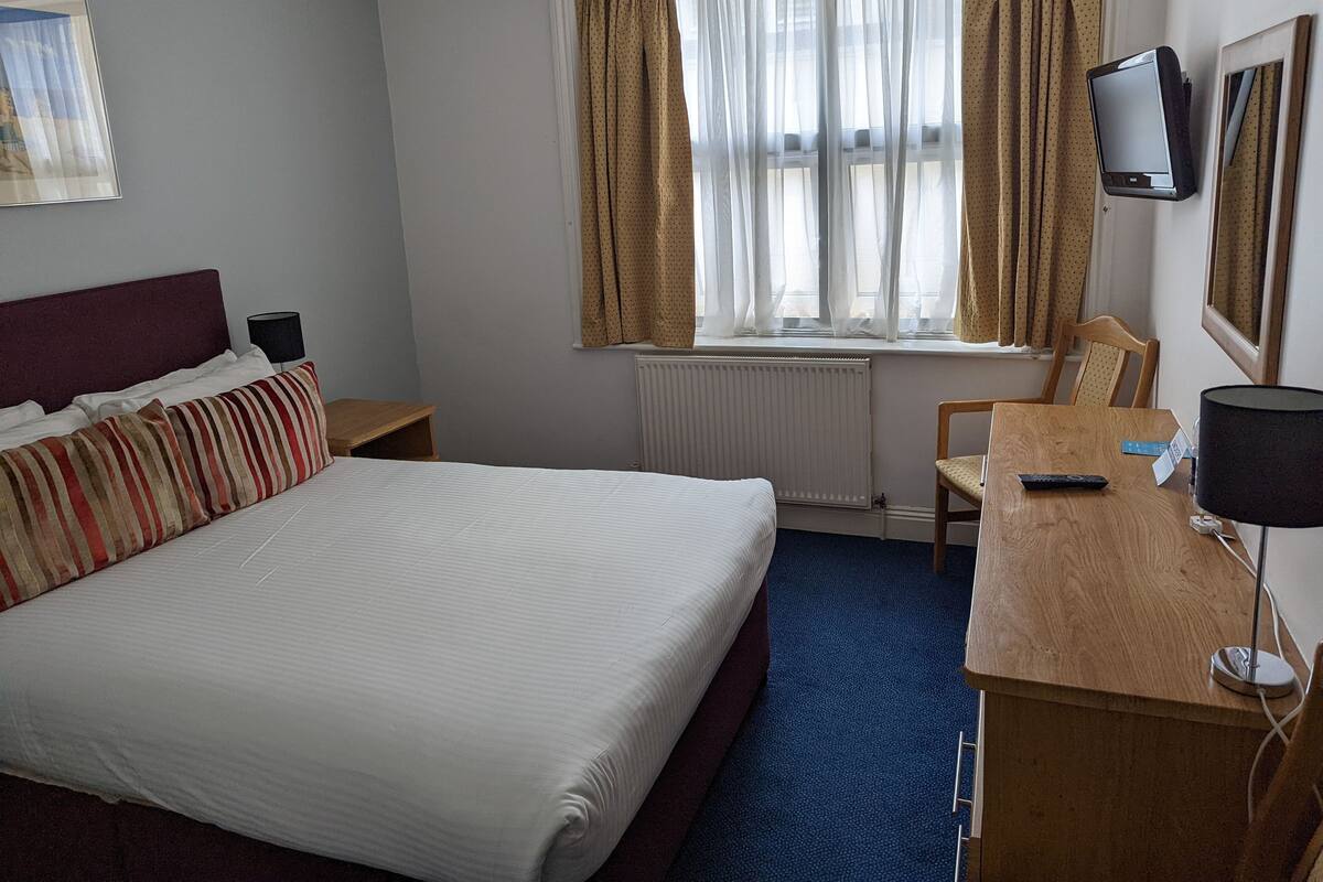 Quarto de hotel com cama, escrivaninha, cadeira, televisão e janela com cortinas.