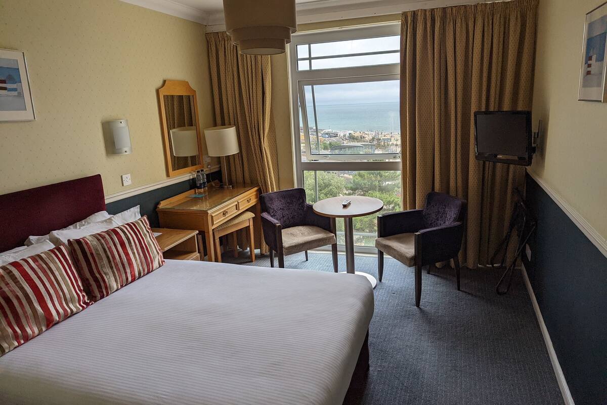 Quarto de hotel com cama, escrivaninha, duas cadeiras, uma mesinha, televisão e uma janela grande com vista.