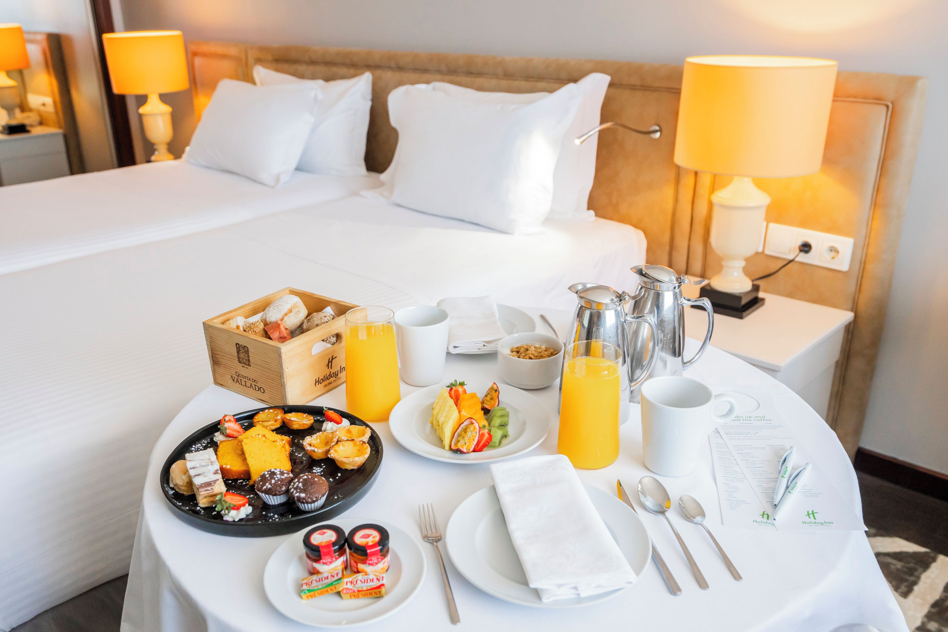 Mesa de café da manhã bem posta, com diversos pães, frutas e bebidas, em quarto de hotel com cama grande e abajures.