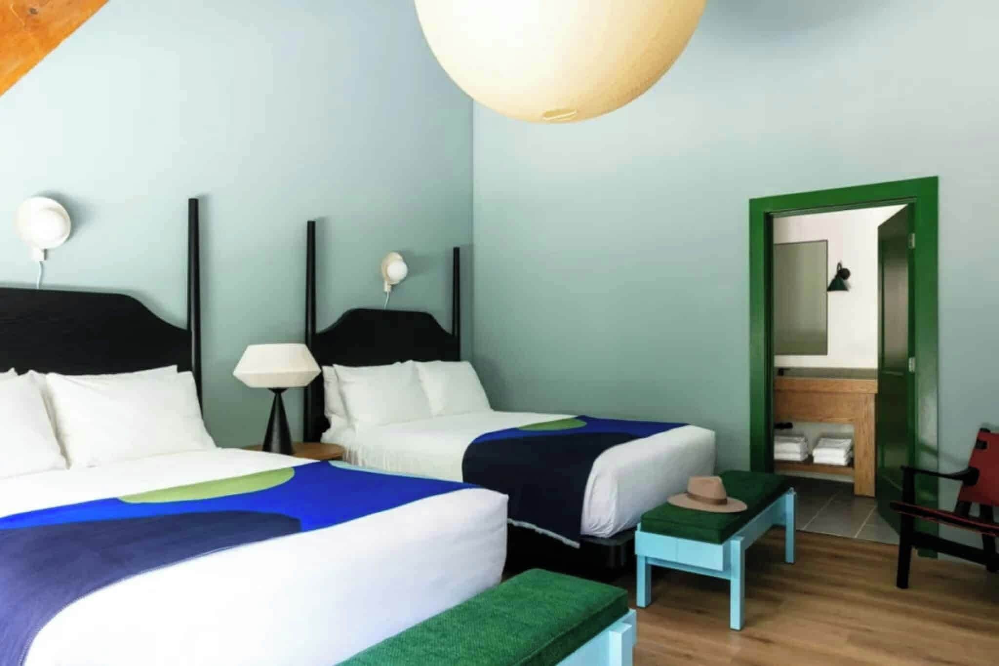 Quarto de hotel com duas camas, mesa de cabeceira, luminária, espelho e um pequeno banco.