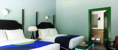 Quarto de hotel com duas camas, mesa de cabeceira, luminária, espelho e um pequeno banco.