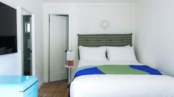 Quarto de hotel com uma cama, mesa de cabeceira, luminária e mala azul.