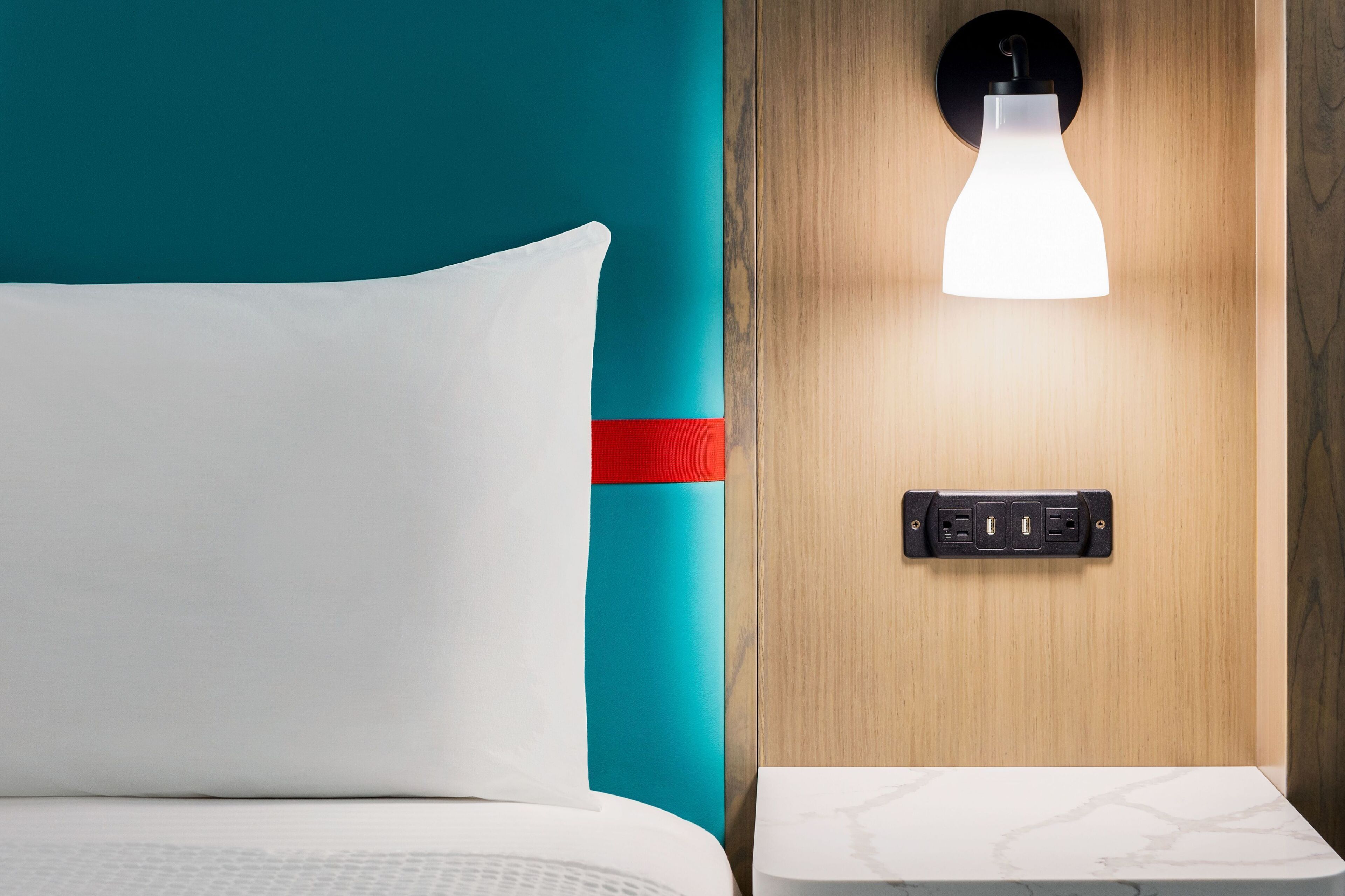Cama com roupa de cama branca, parede verde-água e painel de madeira com luminária e tomadas.