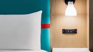 Cama com roupa de cama branca, parede verde-água e painel de madeira com luminária e tomadas.