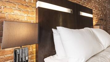 Quarto de hotel com parede de tijolos, cabeceira de madeira escura, roupa de cama branca e uma luminária moderna na mesa de cabeceira.