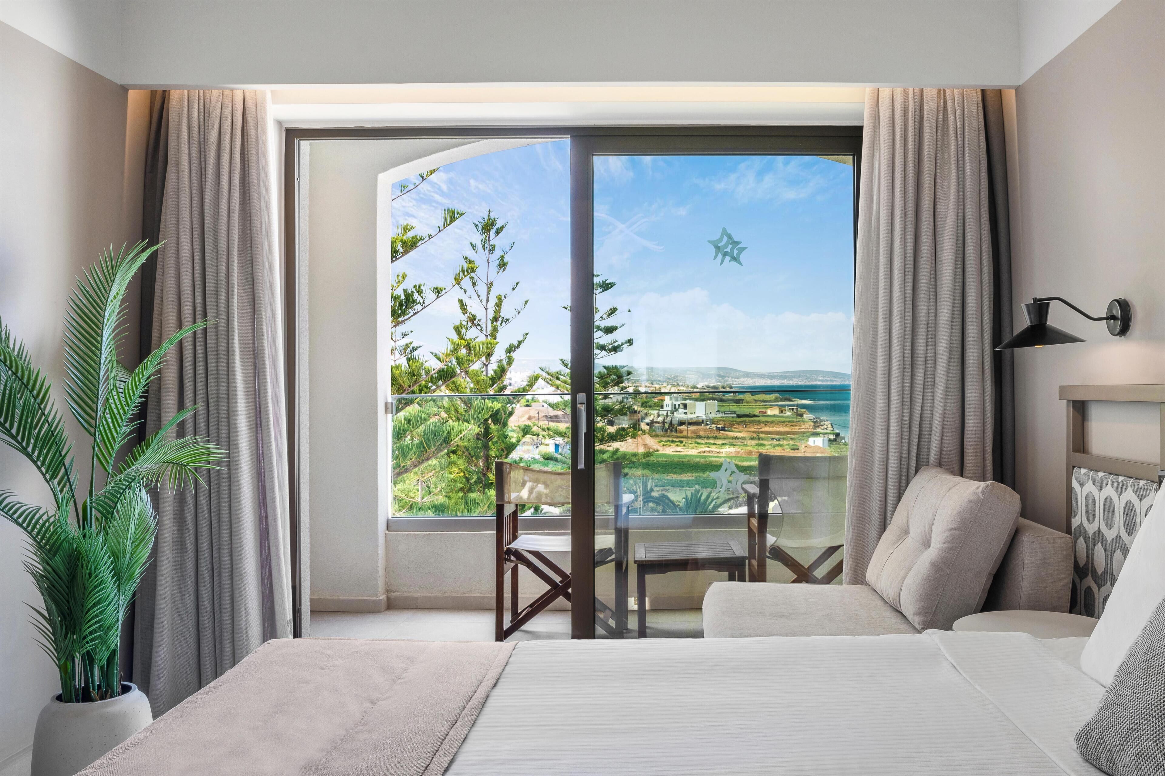 Quarto de hotel com uma grande porta de vidro deslizante que dá para uma varanda com vista para o mar e vegetação.
