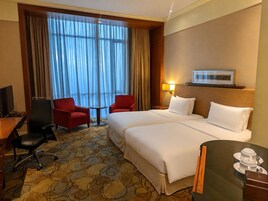 Quarto de hotel com uma cama grande, duas poltronas vermelhas, uma escrivaninha com computador e uma janela com cortinas.