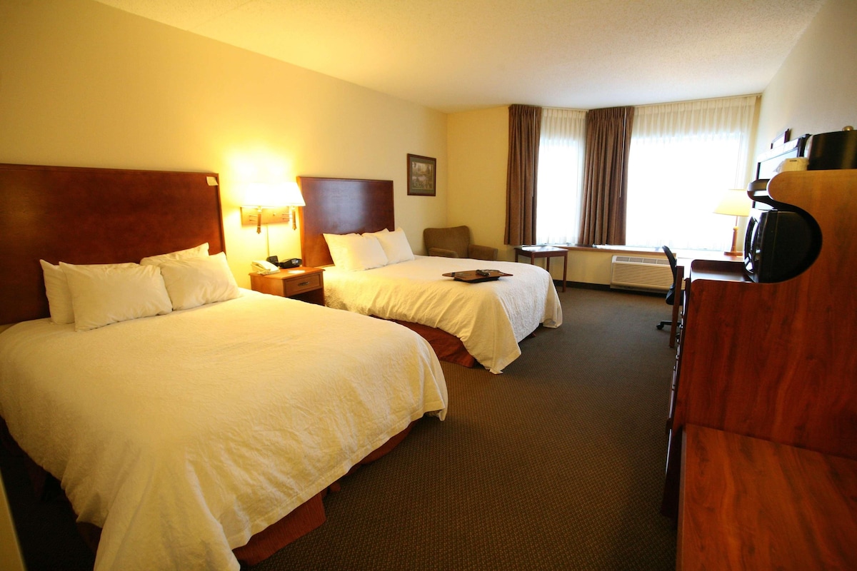 Quarto de hotel com duas camas, uma escrivaninha, uma cadeira e uma televisão.