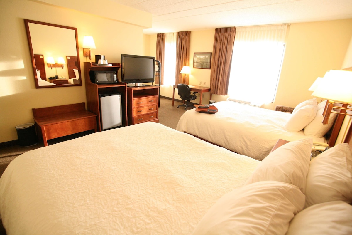 Quarto de hotel com duas camas, uma escrivaninha, uma televisão e um espelho.