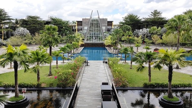 Área de piscina com um caminho central, cercada por palmeiras e um edifício com fachada de vidro.