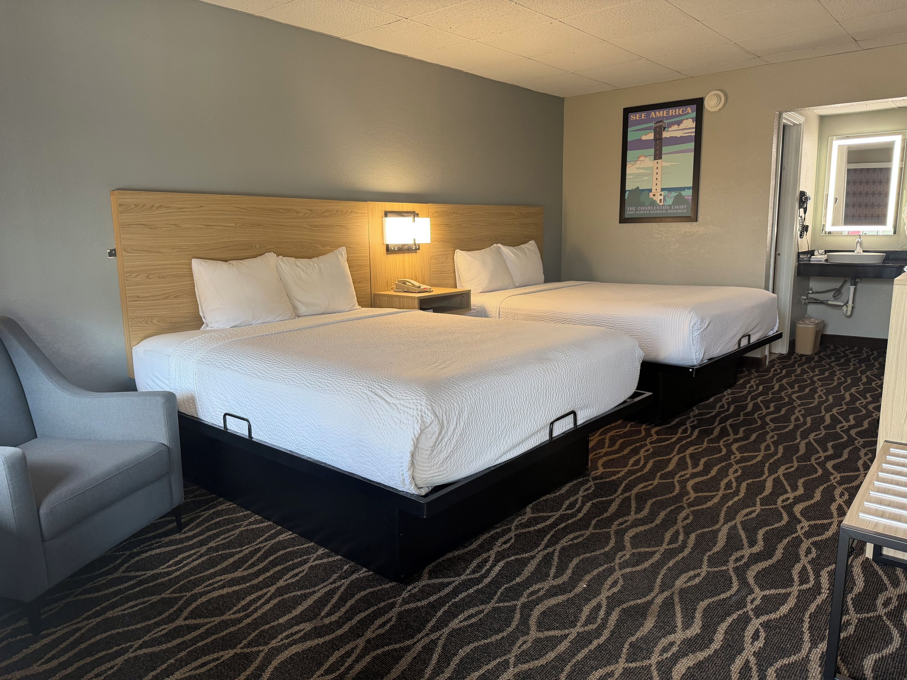 Quarto de hotel com duas camas, uma escrivaninha, uma cadeira, um banheiro e um pôster na parede.