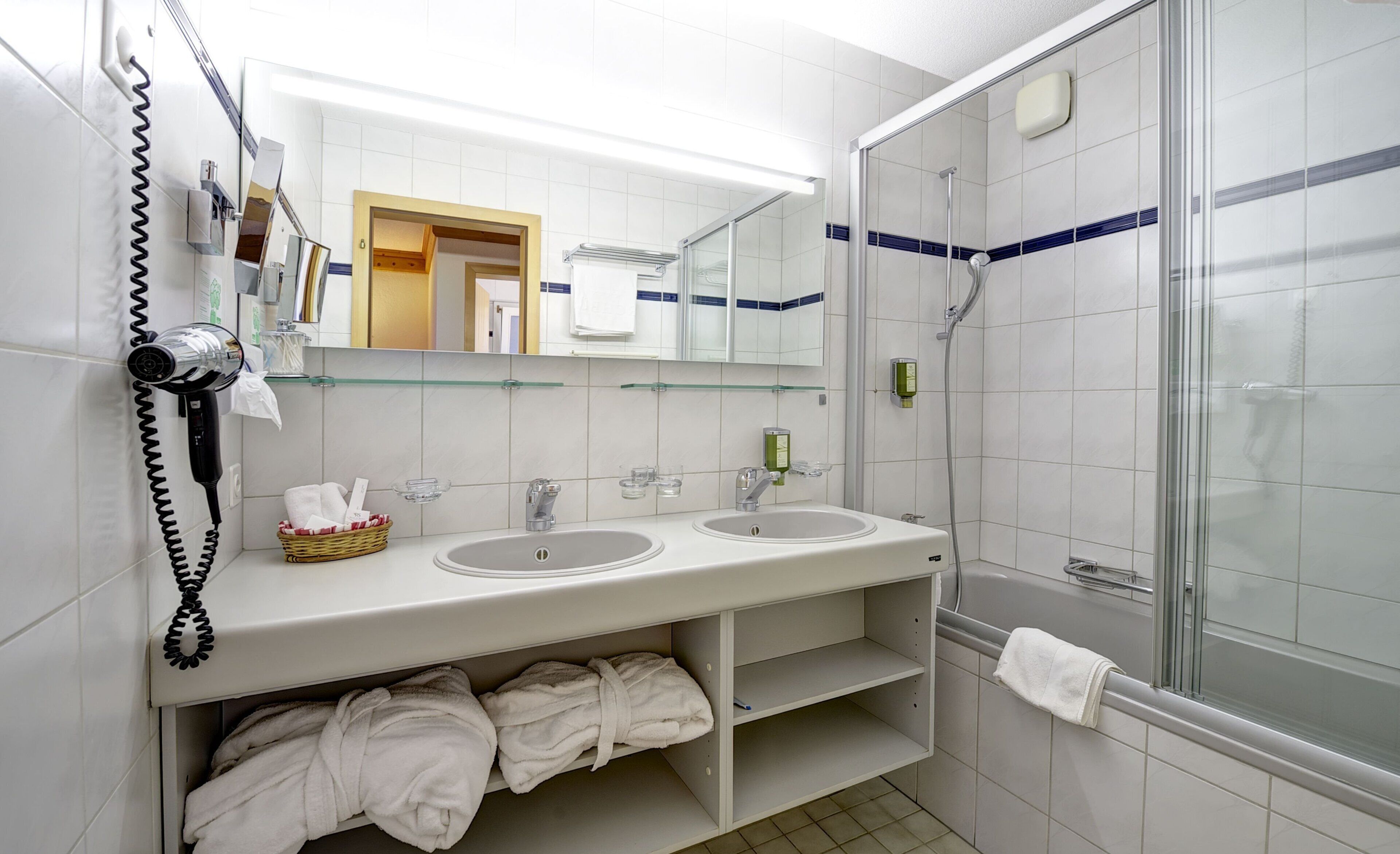 Casa de banho com duas pias, cabine de duche em vidro, secador de cabelo e espelho.