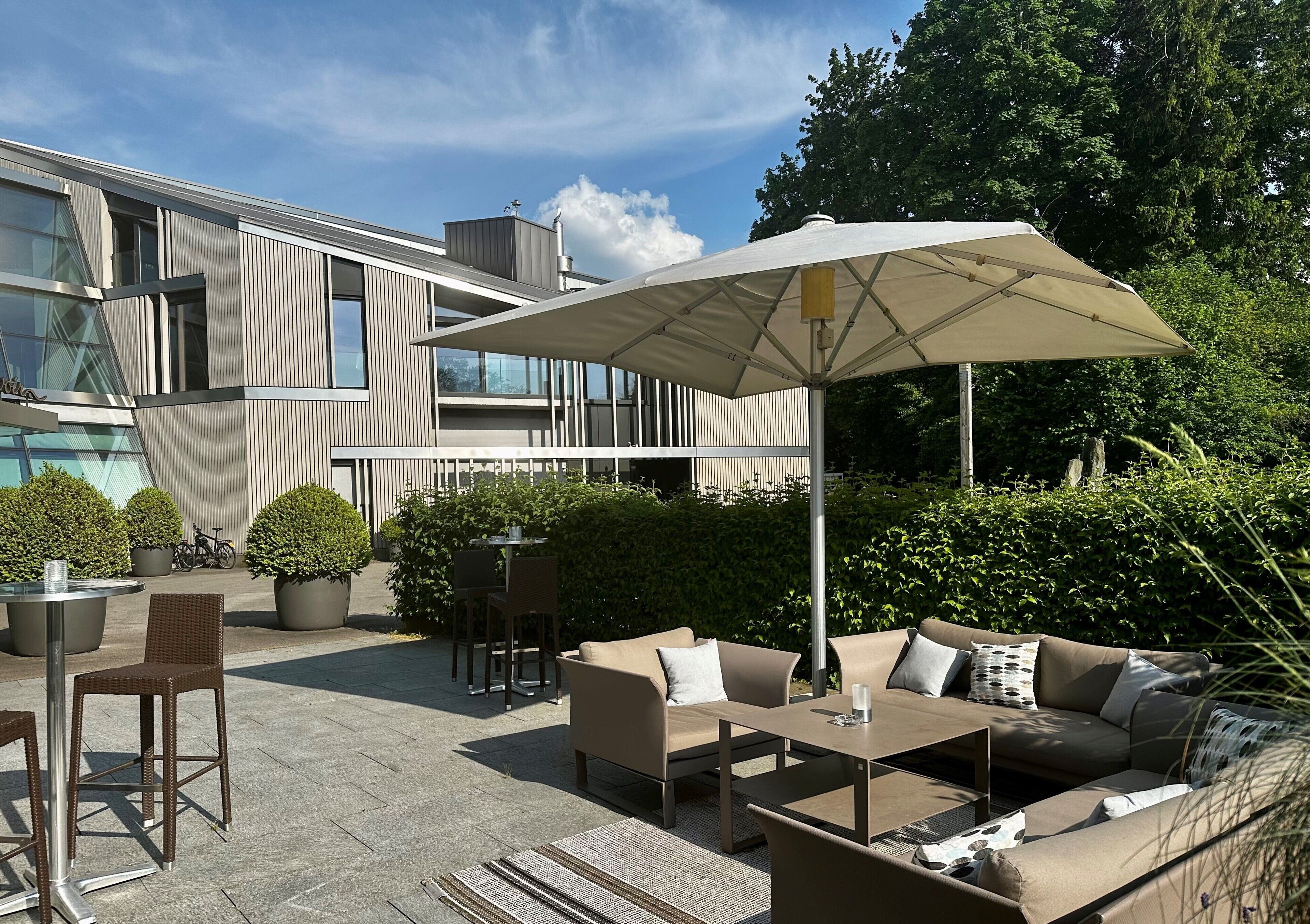 Uma área de terraço ao ar livre com móveis modernos, incluindo um sofá, cadeiras e uma mesa, sob um grande guarda-sol.