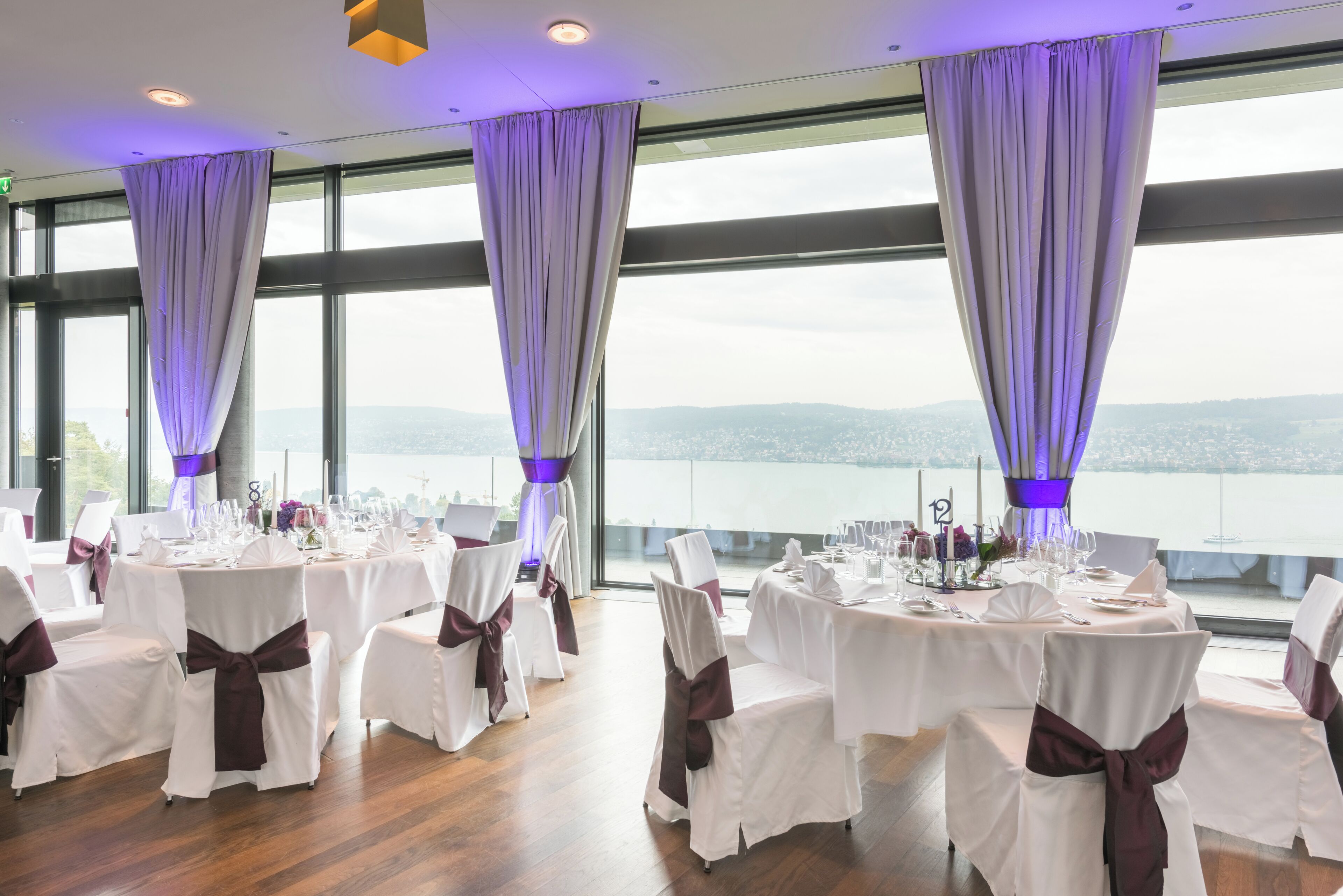 Uma sala de banquetes com mesas redondas, toalhas brancas e cadeiras com fitas escuras, com vista para um corpo de água.
