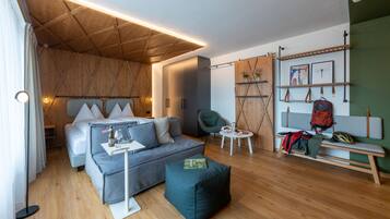 Quarto moderno e bem iluminado com uma cama, um sofá cinza, uma mesa pequena com uma garrafa e um copo, e um banquinho verde.