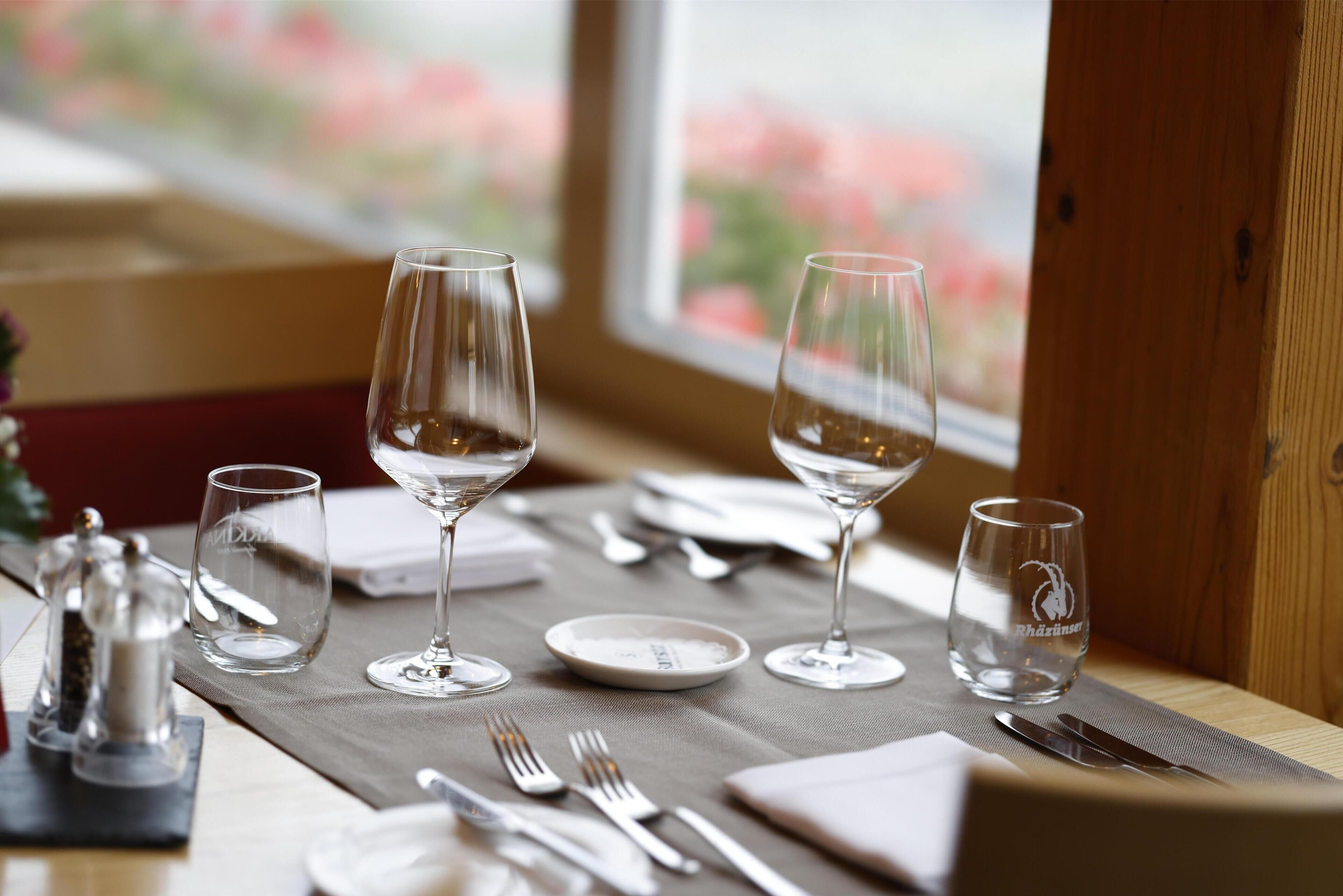 Mesa posta com taças de vinho, talheres e pratos, com vista para flores através de uma janela.